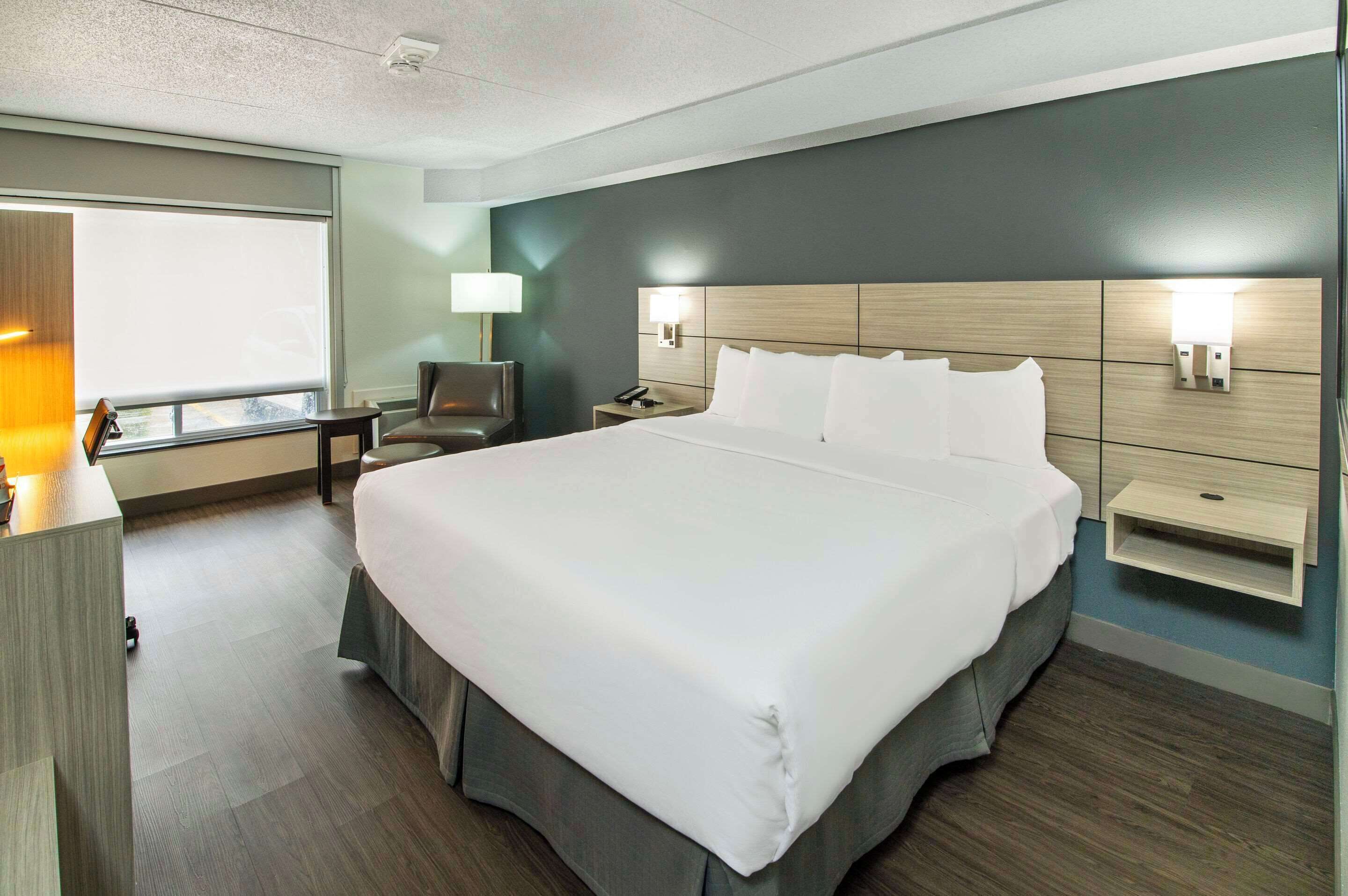 Quarto de hotel com uma cama grande, uma escrivaninha com luminária, uma cadeira e uma mesa de cabeceira com telefone.