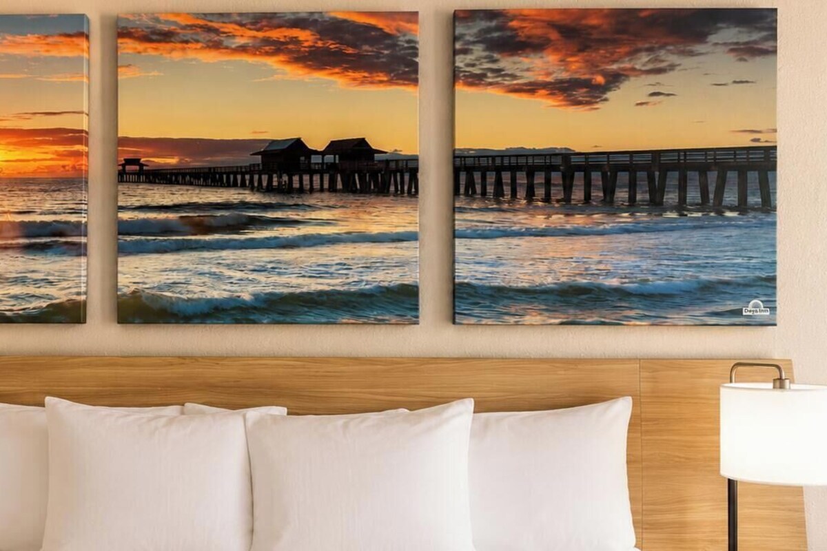 Quarto de hotel com cabeceira de madeira, almofadas brancas e uma obra de arte na parede retratando um pôr do sol sobre o oceano com um píer.