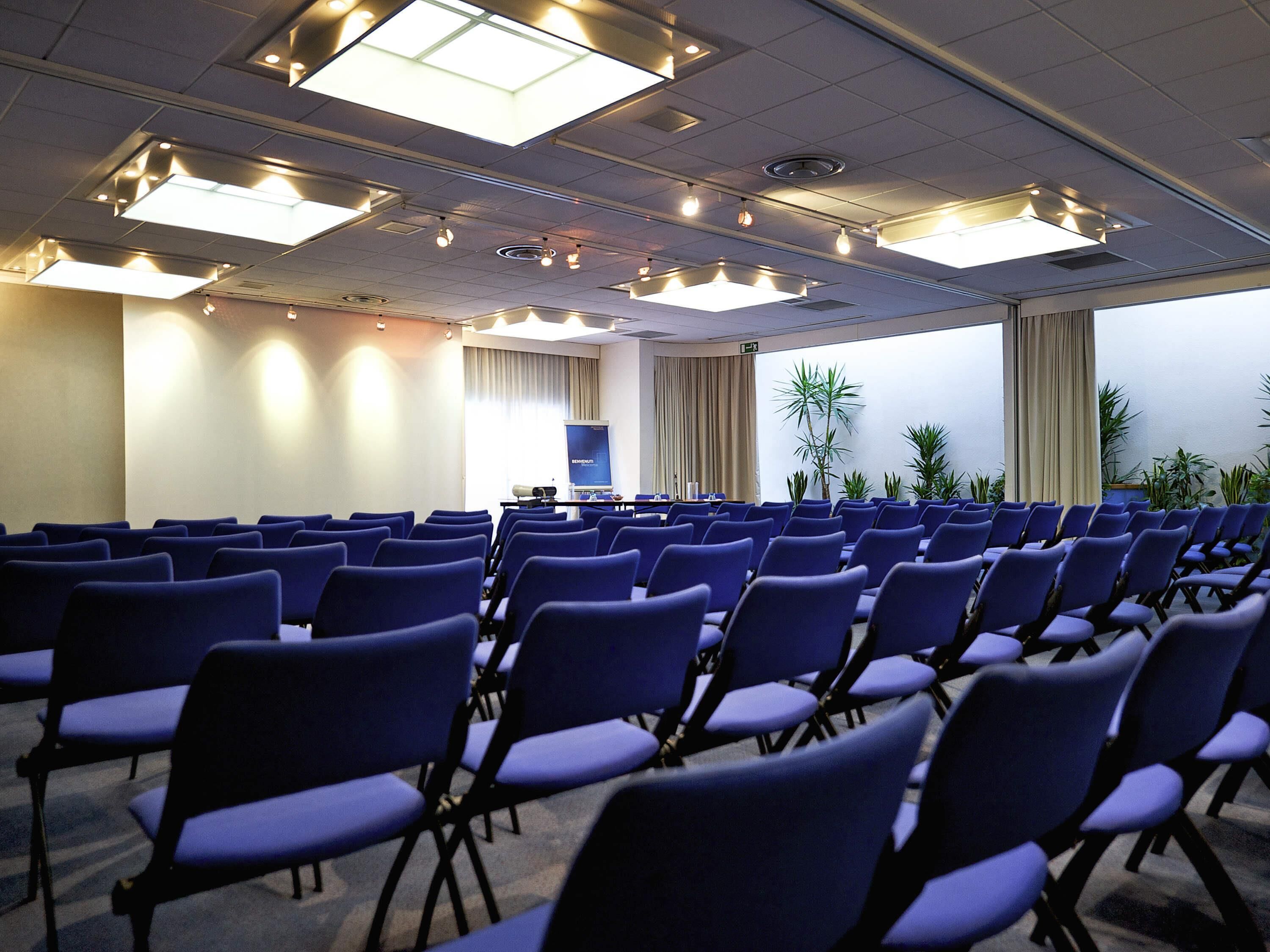 Sala de conferências com fileiras de cadeiras, uma tela de projeção e grandes janelas.