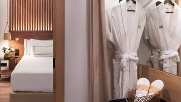 Quarto de hotel com cama, mesa de cabeceira, abajur, espelho e um suporte com roupões e chinelos.