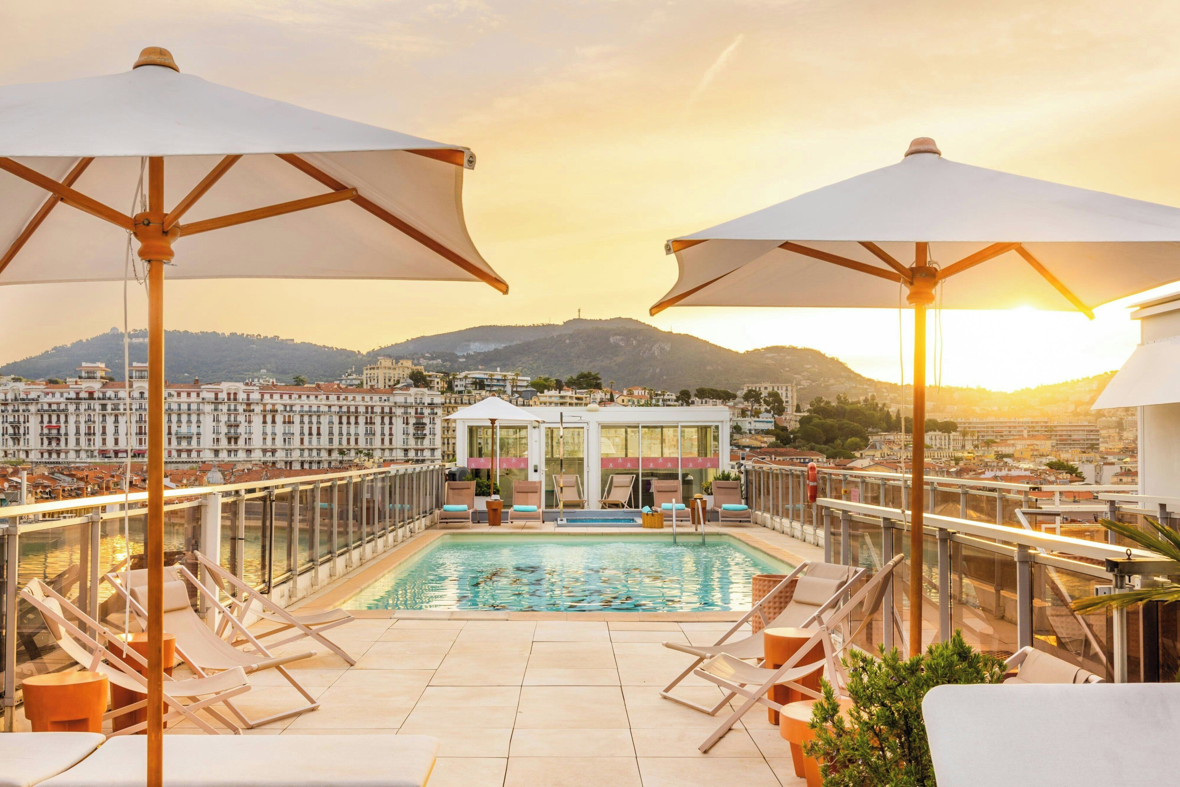 Área de piscina no terraço com espreguiçadeiras e guarda-sóis, com vista para um horizonte urbano com edifícios e colinas.