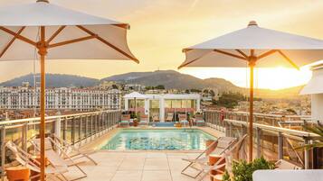 Área de piscina no terraço com espreguiçadeiras e guarda-sóis, com vista para um horizonte urbano com edifícios e colinas.