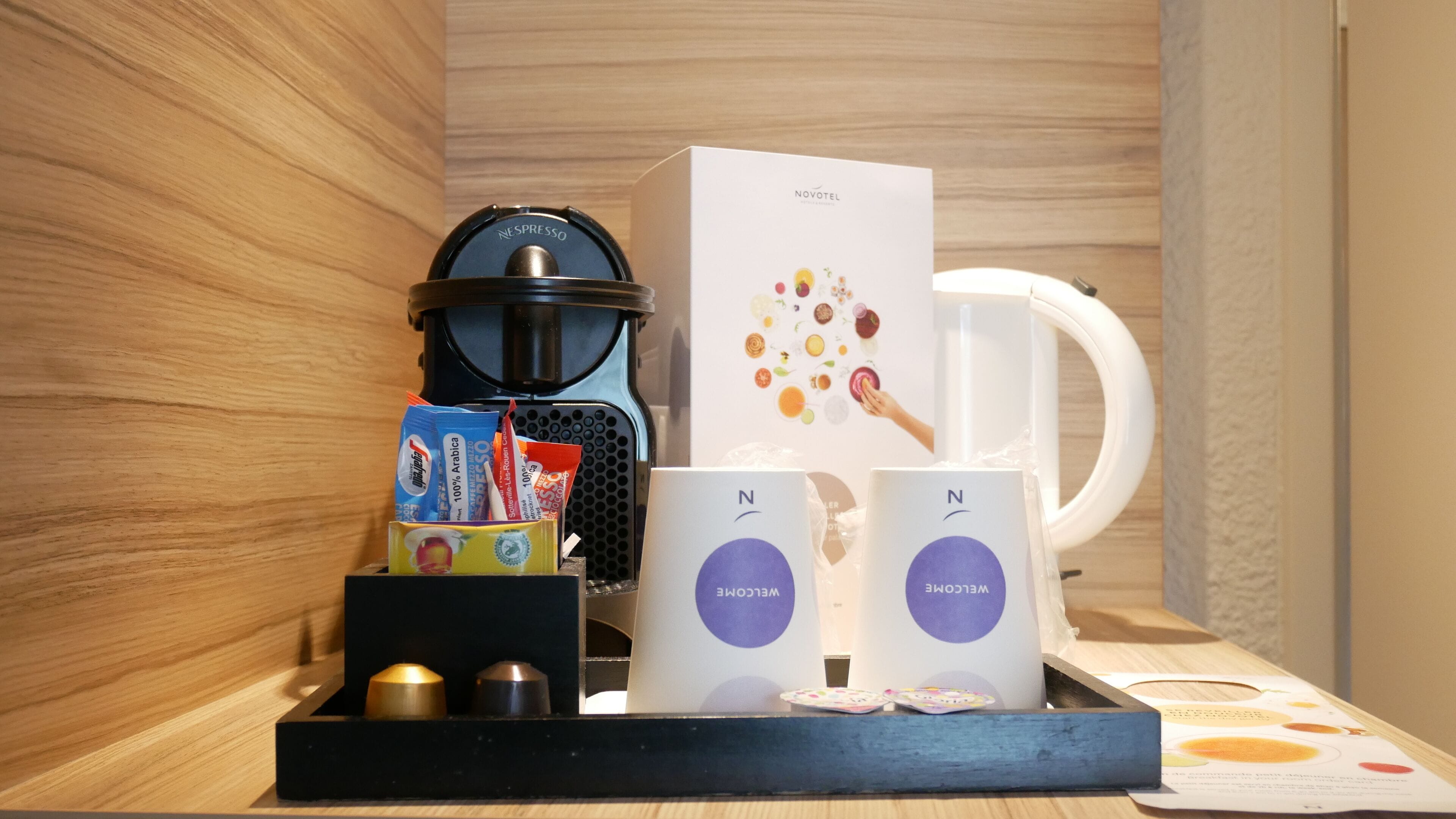 Uma estação de café com uma cafeteira, uma caixa com a inscrição 'NOVOTEL' e duas canecas com a palavra 'BEM-VINDO'.