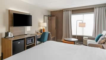 Quarto de hotel com cama, escrivaninha com televisão de tela plana, micro-ondas, mini-geladeira, cadeira, mesa redonda, luminária e uma janela grande com cortinas.