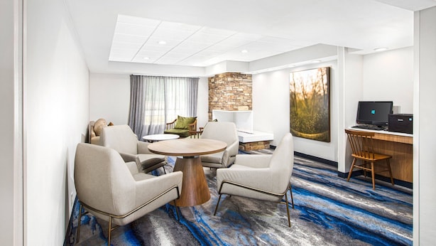 Quarto de hotel moderno com uma mesa redonda, quatro poltronas, uma escrivaninha com um laptop e um grande quadro abstrato na parede.