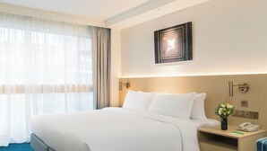 Quarto de hotel com uma cama grande, uma mesa de cabeceira com telefone e um vaso de flores, uma luminária na parede e uma janela com cortinas leves.