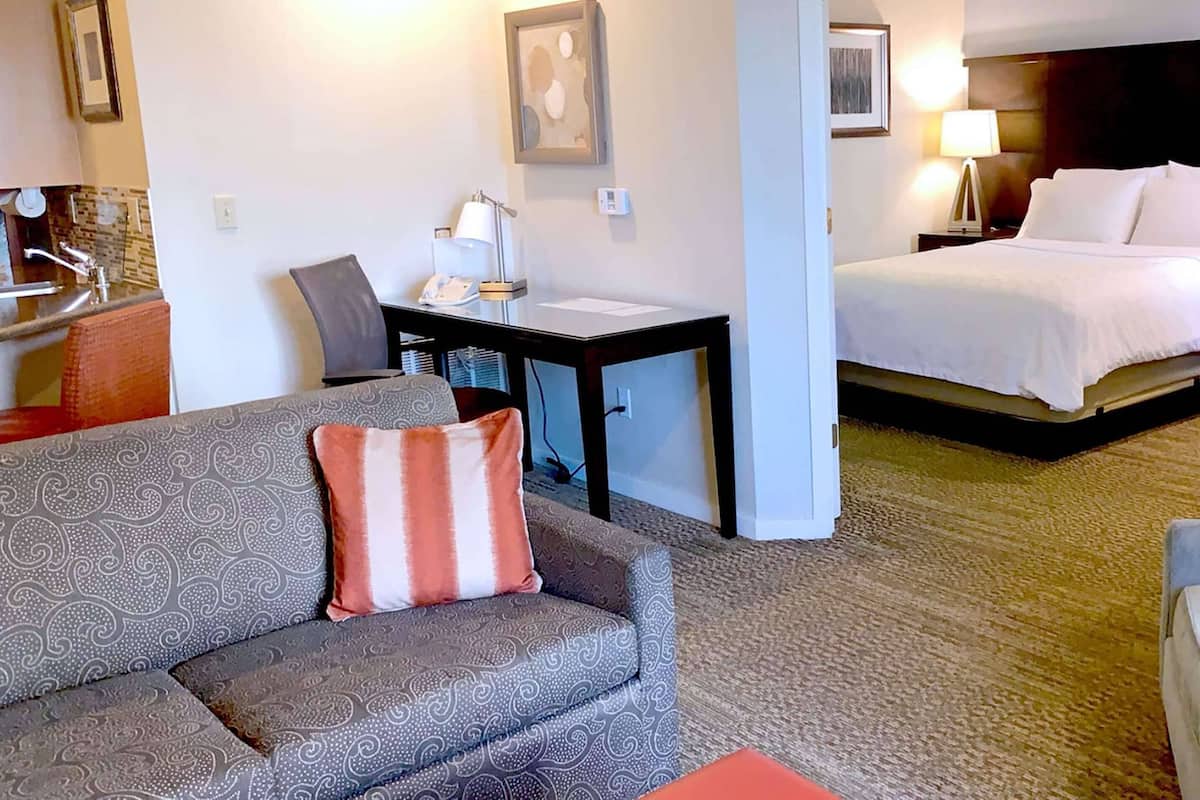 Quarto de hotel com sofá, poltrona, escrivaninha, mesa de cabeceira, luminária e cama.