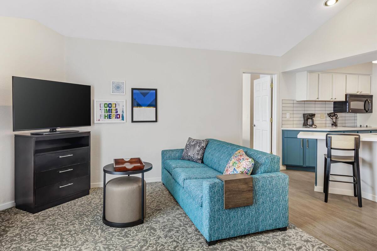 Uma sala de estar moderna com sofá, TV e uma pequena mesa lateral. Uma área de cozinha é visível, com armários e micro-ondas.