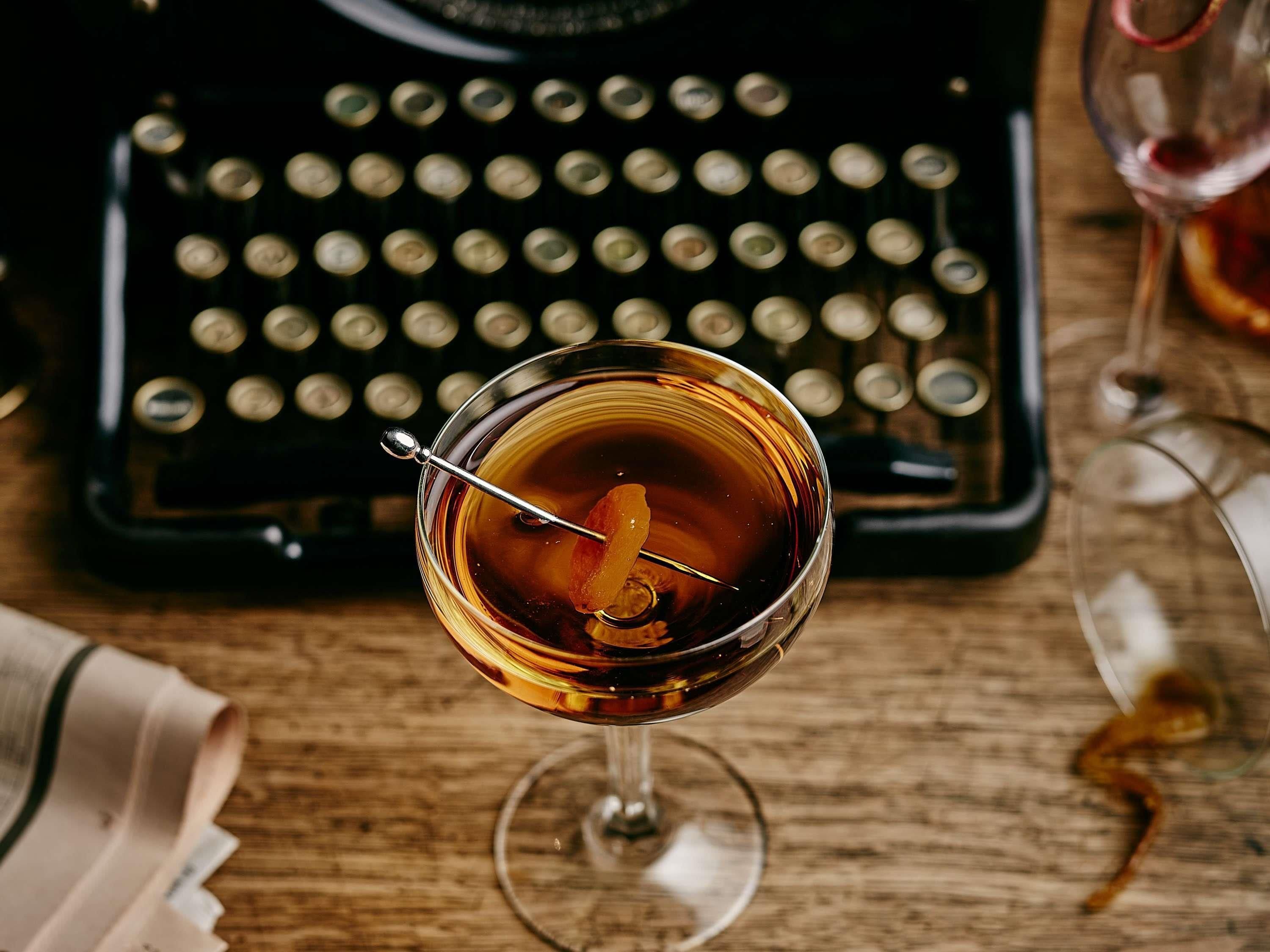 Um copo com bebida, uma máquina de escrever e um livro sobre uma superfície de madeira.