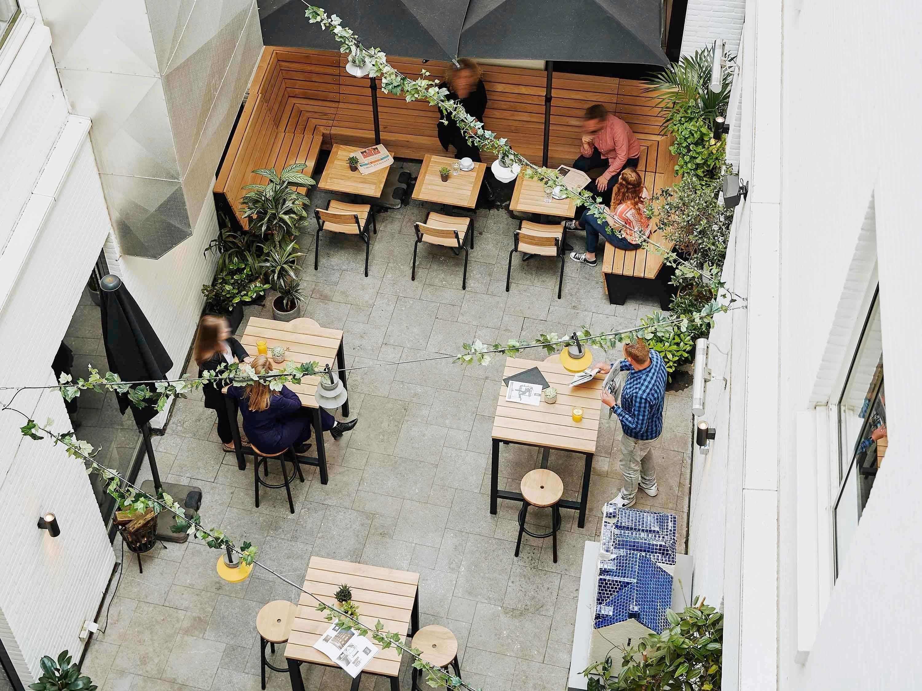 Um café ao ar livre com mesas e cadeiras de madeira, rodeado por plantas em vasos e um espaço com bancos de madeira.