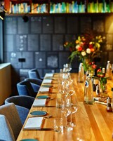 Mesa de madeira comprida posta para refeição, com cadeiras, copos de vinho e garrafas.