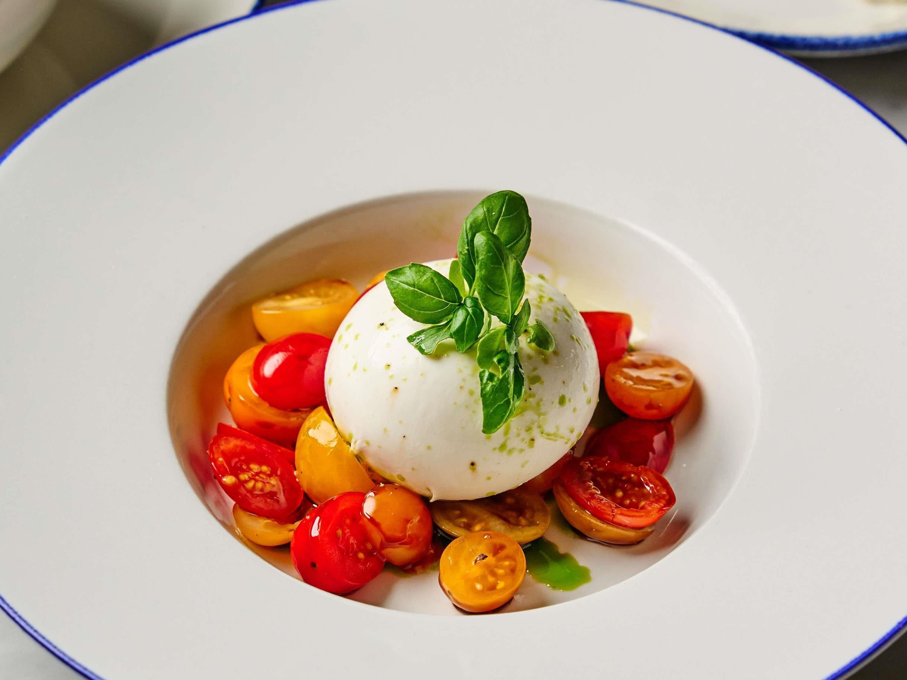 Um prato branco com uma esfera branca no centro, rodeado por metades de tomate cereja e pimentões amarelos, decorado com folhas verdes.