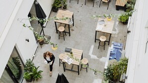 Um pátio externo com várias mesas e cadeiras de madeira, cercado por vasos de plantas e guarda-sóis.