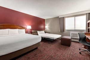 Quarto de hotel com uma cama grande, uma cama menor, uma escrivaninha, uma cadeira, um abajur e uma janela com cortinas.