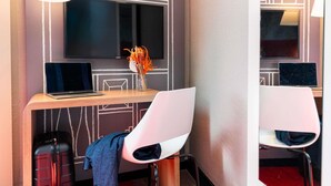 Quarto de hotel moderno com uma televisão de tela plana grande, dois laptops, uma mesa com um vaso de flores e duas cadeiras brancas.