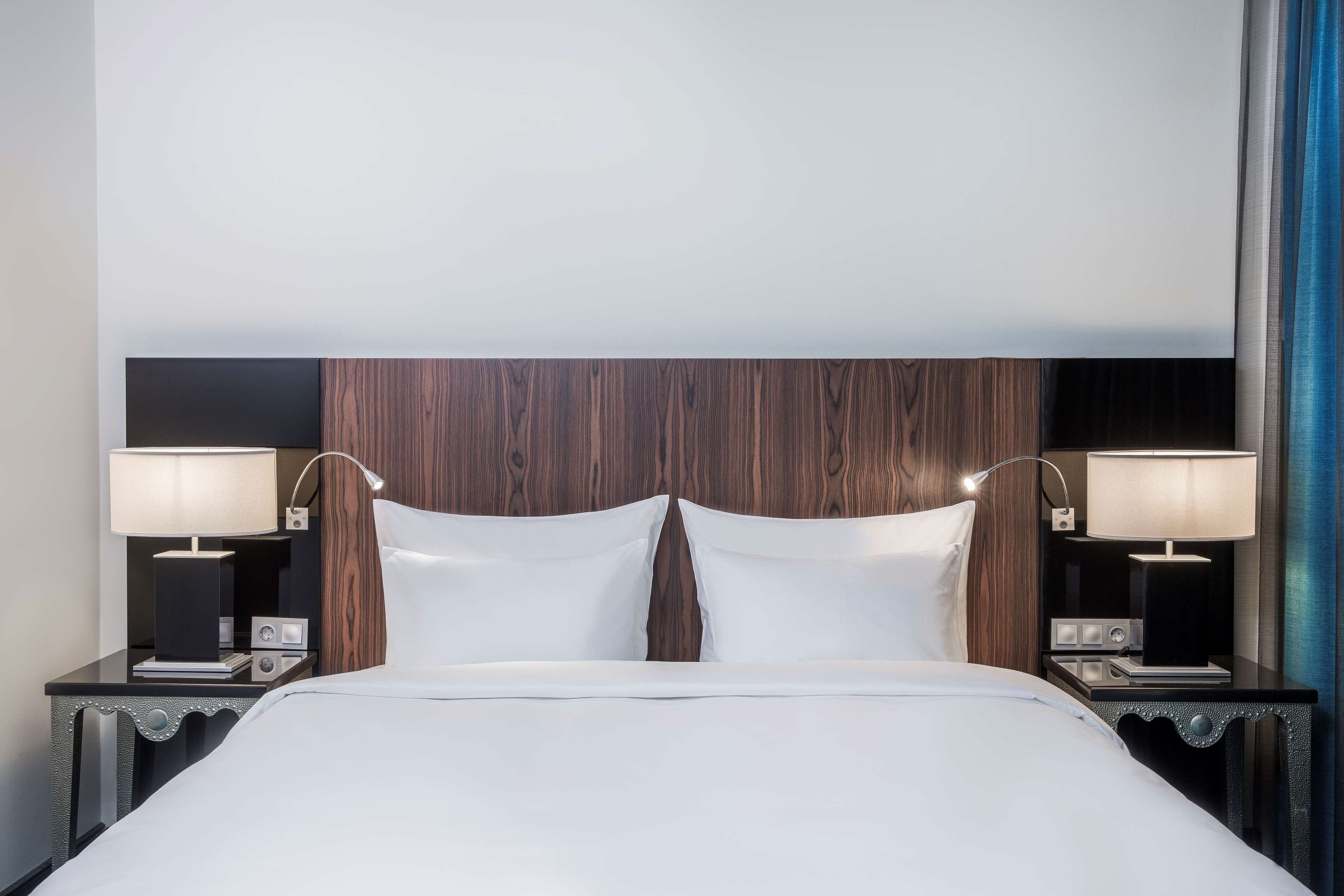 Cama bem arrumada com lençóis brancos, com duas mesas de cabeceira com luminárias, ao fundo um painel de cama de madeira e uma parede lisa.