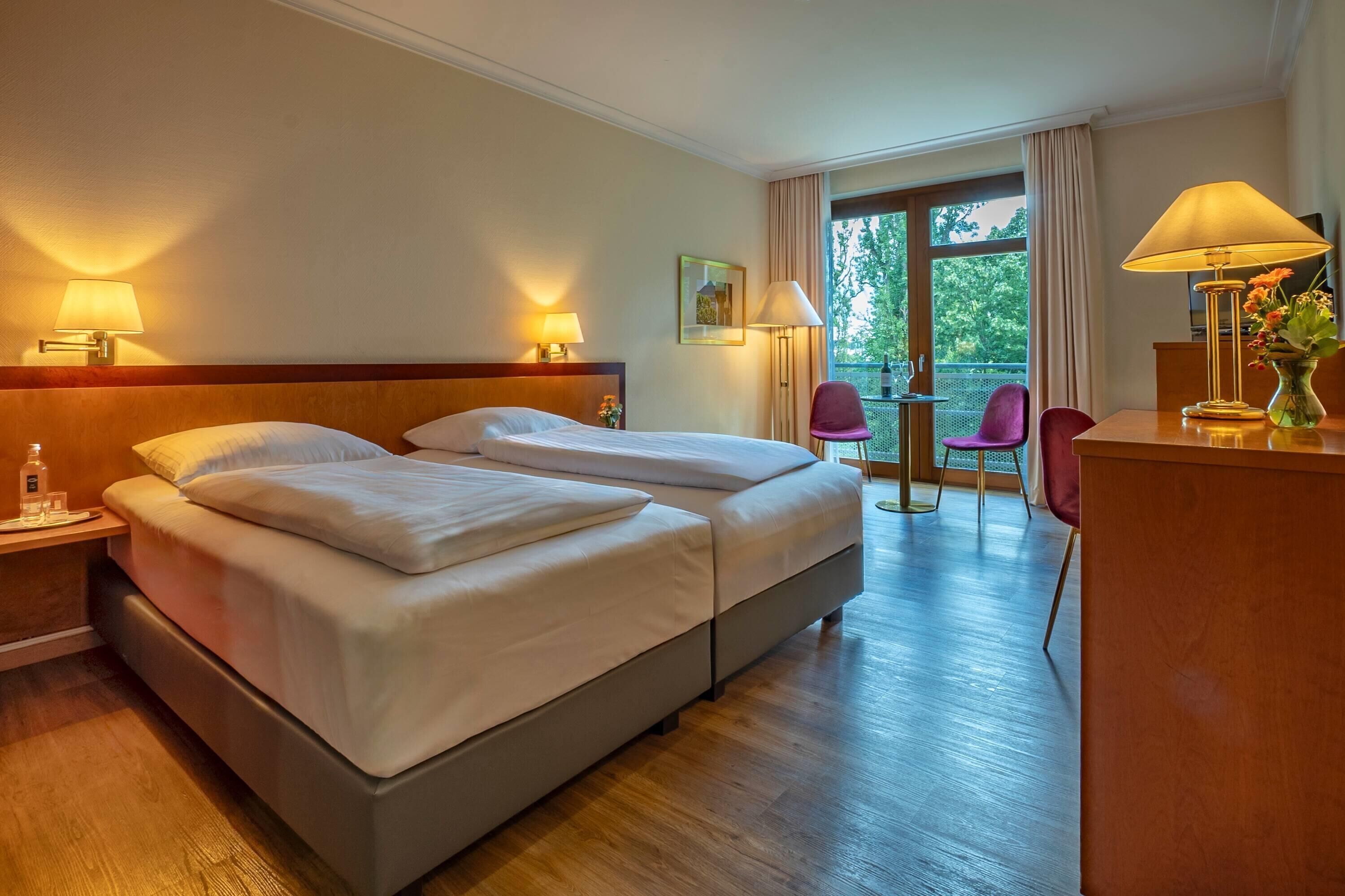 Quarto de hotel com duas camas, uma mesa, uma cadeira e vista para o verde lá fora.