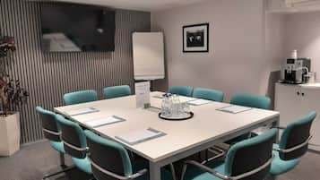 Uma sala de conferências com uma mesa branca, cadeiras verde-água, uma TV fixada na parede, um quadro branco e uma máquina de café.
