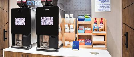 Uma estação de café com duas máquinas de café, um aviso indicando o horário do café da manhã gratuito e diversos produtos de café e chá nas prateleiras.