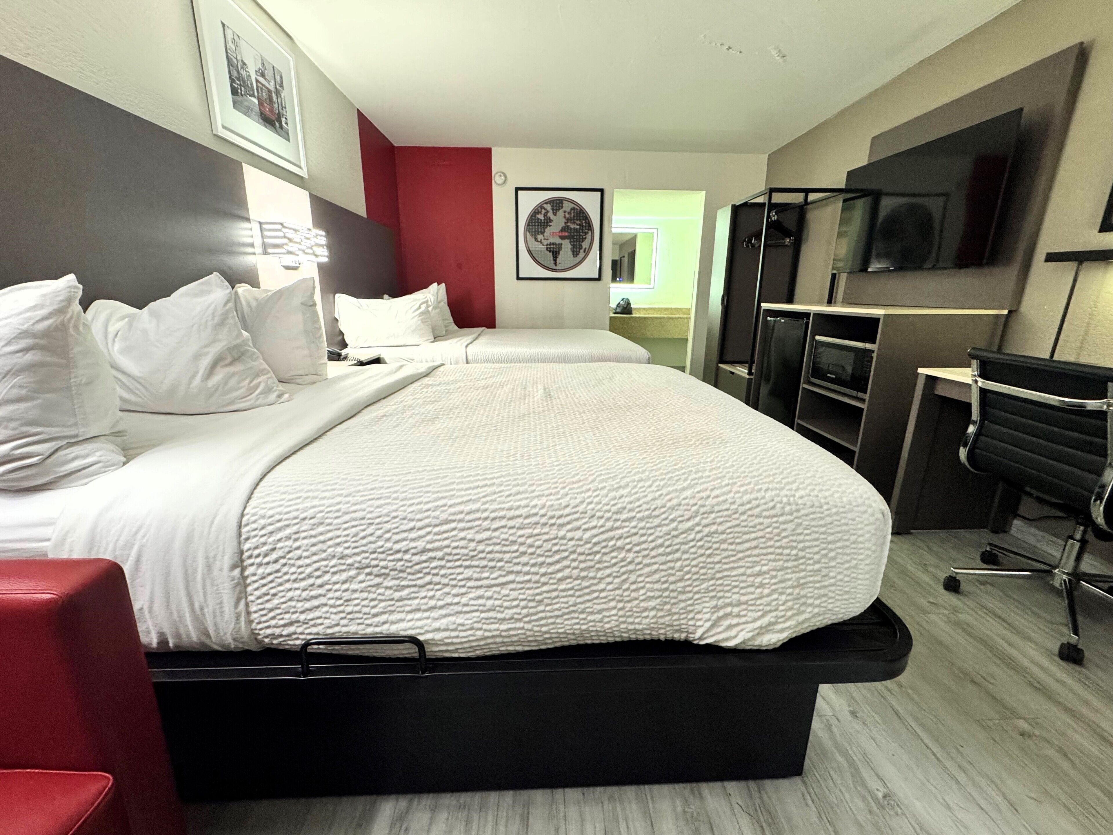 Quarto de hotel com duas camas, uma mesa, uma cadeira, uma televisão e uma parede de destaque vermelha.