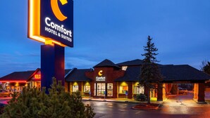Um Comfort Hotel & Suites com uma placa iluminada bem visível, um estacionamento e fachada do prédio bem iluminada.
