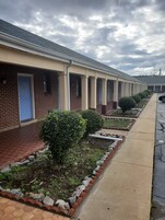 Fileira de quartos de motel com fachadas de tijolo, calçada de concreto e pequenas áreas ajardinadas com arbustos.