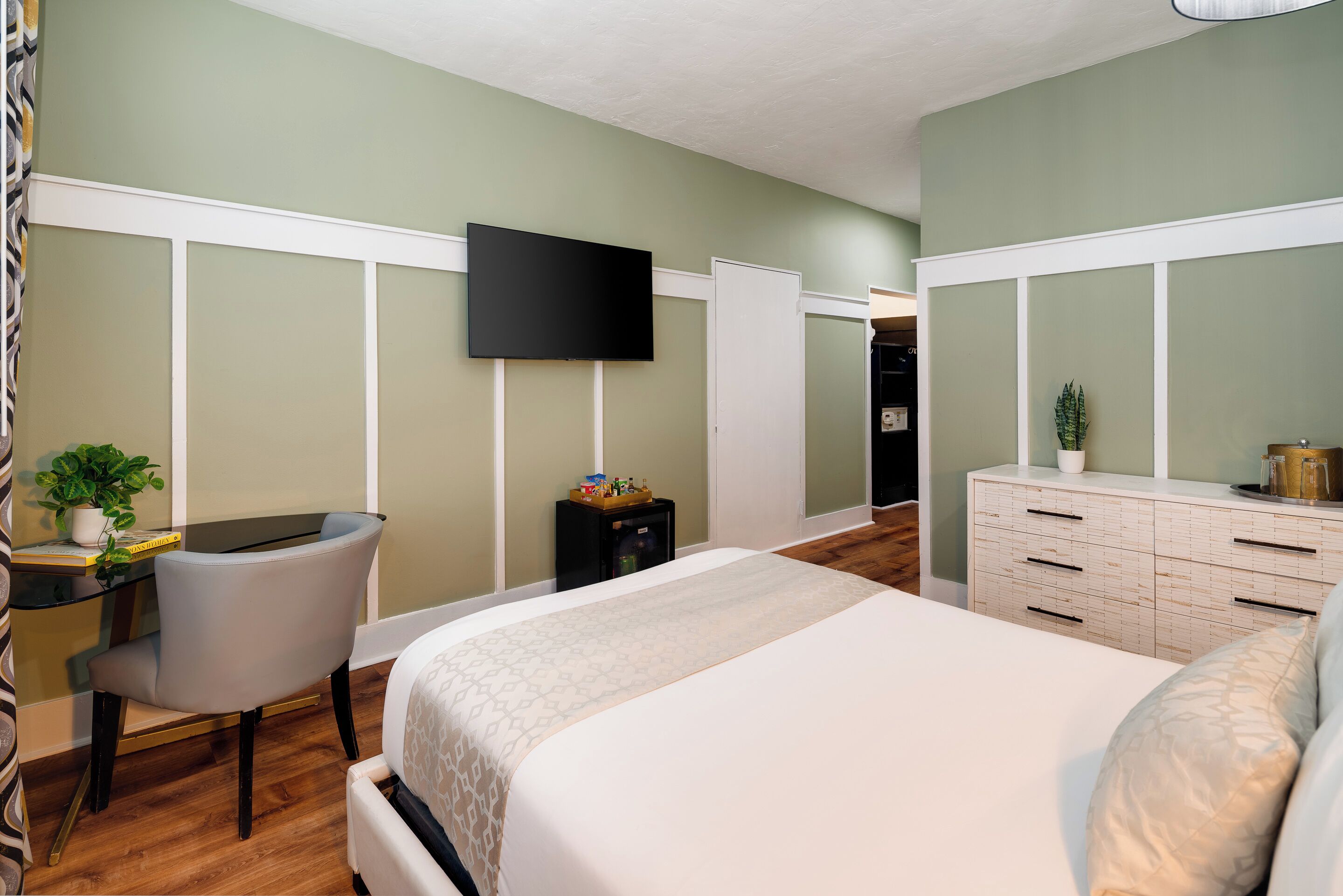 Quarto com cama, escrivaninha, cadeira, cômoda e TV fixada na parede.