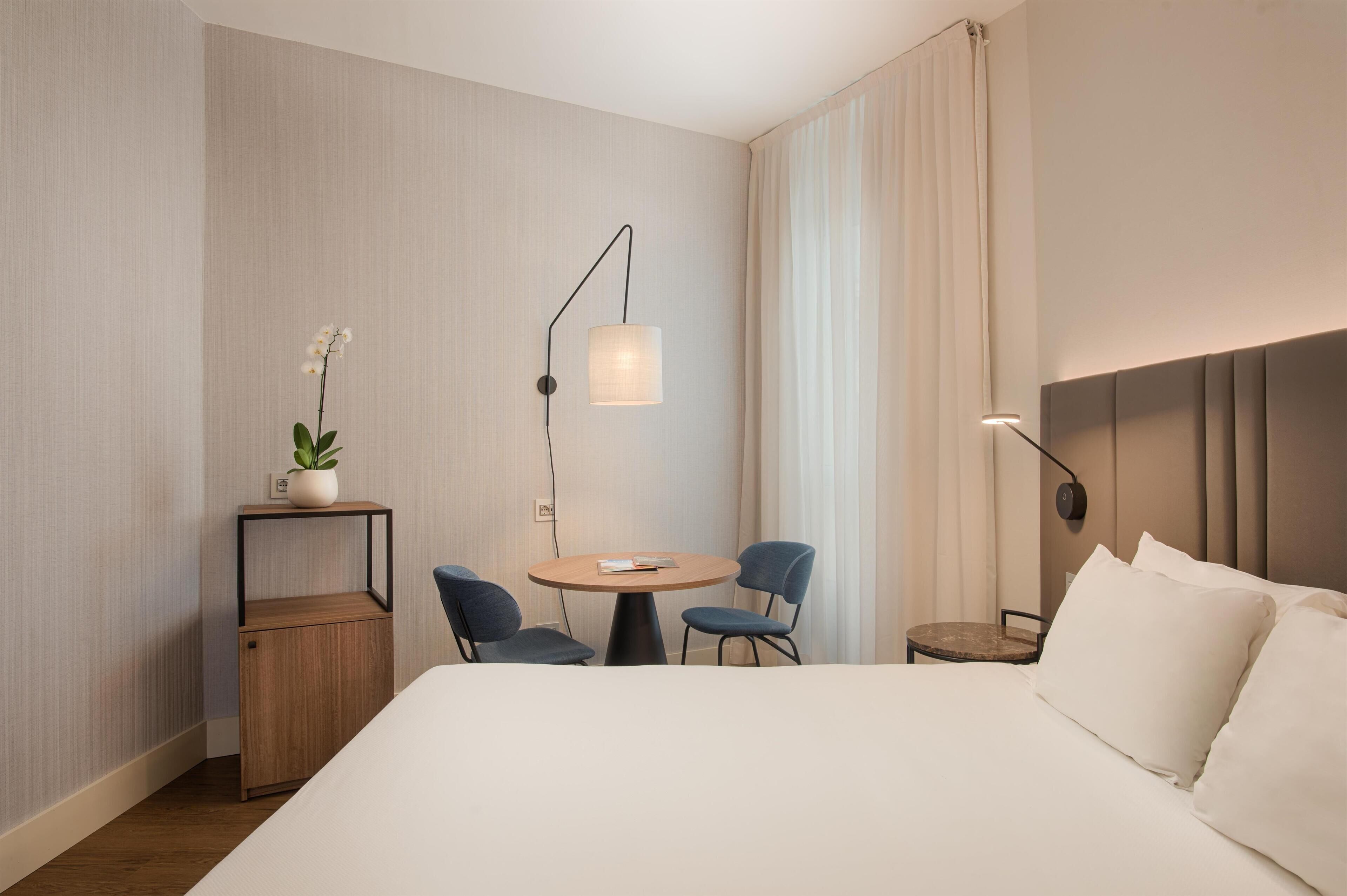 Quarto de hotel com uma cama, uma mesa redonda com cadeiras, uma pequena escrivaninha com abajur e um vaso de planta.