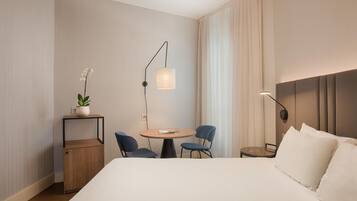 Quarto de hotel com uma cama, uma mesa redonda com cadeiras, uma pequena escrivaninha com abajur e um vaso de planta.
