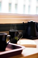 Um cantinho com café, incluindo uma caneca preta, um sachê de chá e um livro sobre uma mesa de madeira perto de uma janela.