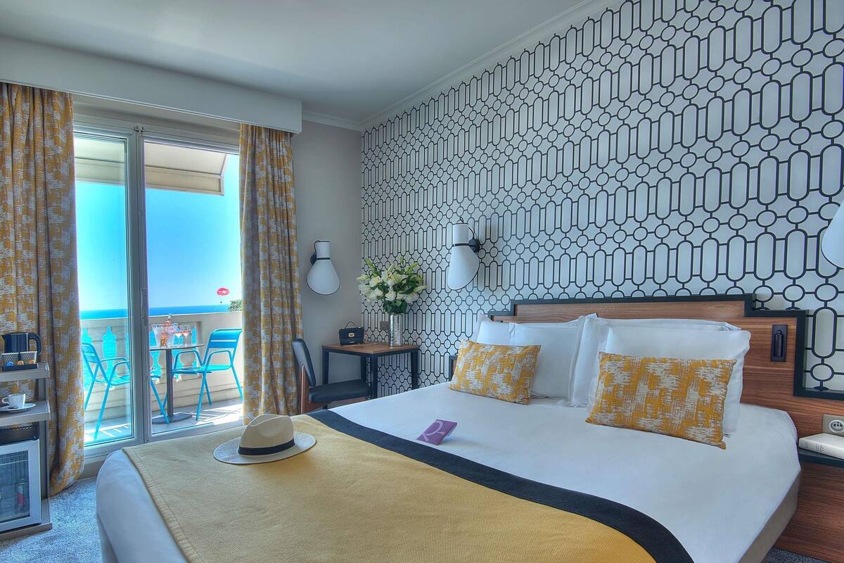 Quarto de hotel com cama, escrivaninha, cadeira, mesa com cadeira, janela com cortinas e parede estampada.
