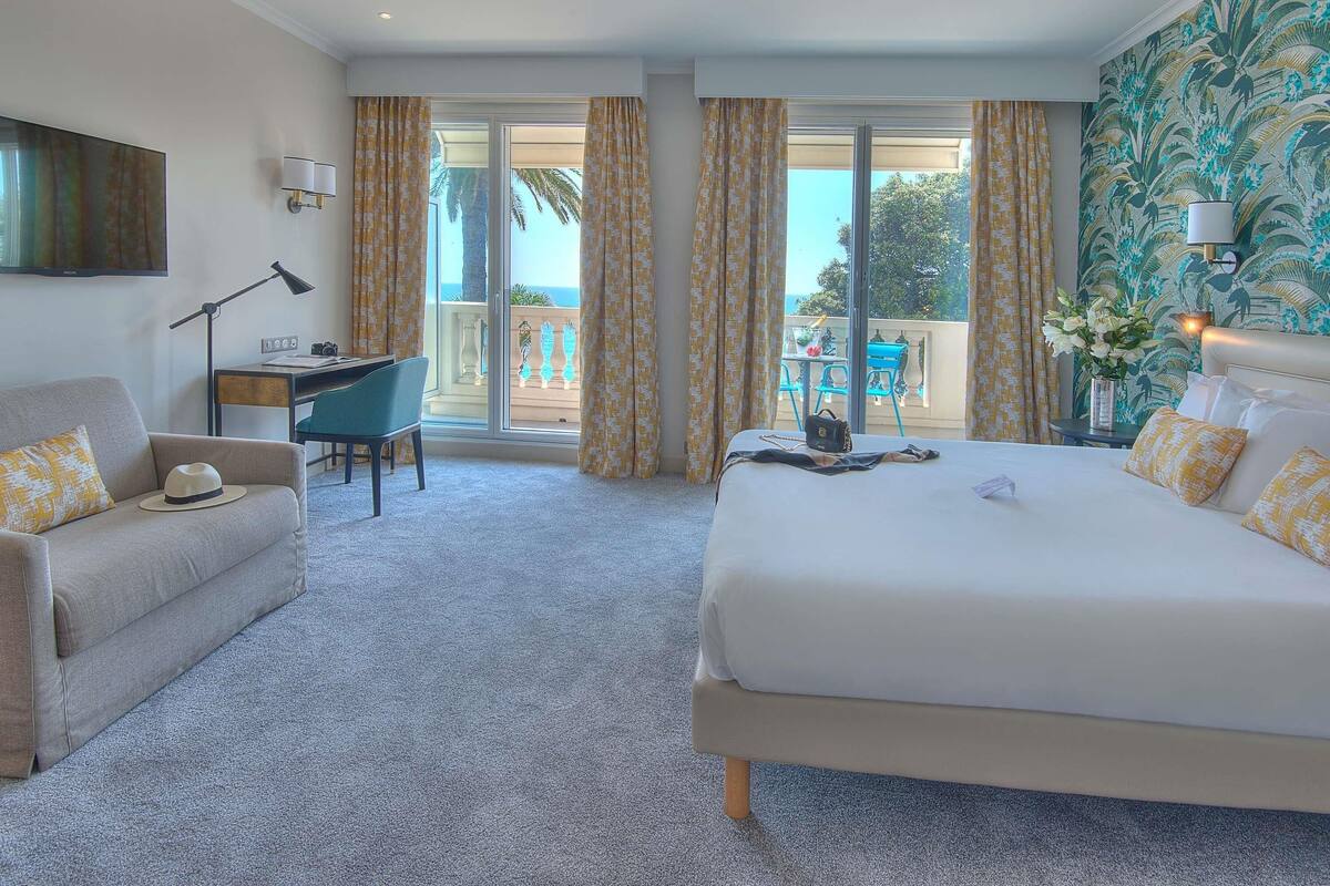Quarto de hotel com cama, escrivaninha, poltrona, sofá e varanda com vista.