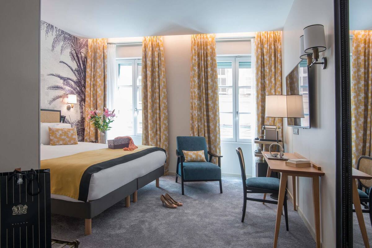 Quarto de hotel com cama, duas cadeiras, escrivaninha, abajur e uma janela grande com cortinas.