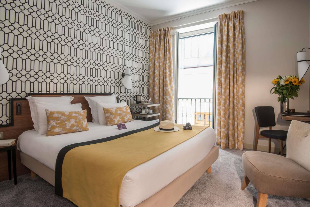 Quarto de hotel com cama grande, escrivaninha, cadeira, parede estampada e vista pela janela.