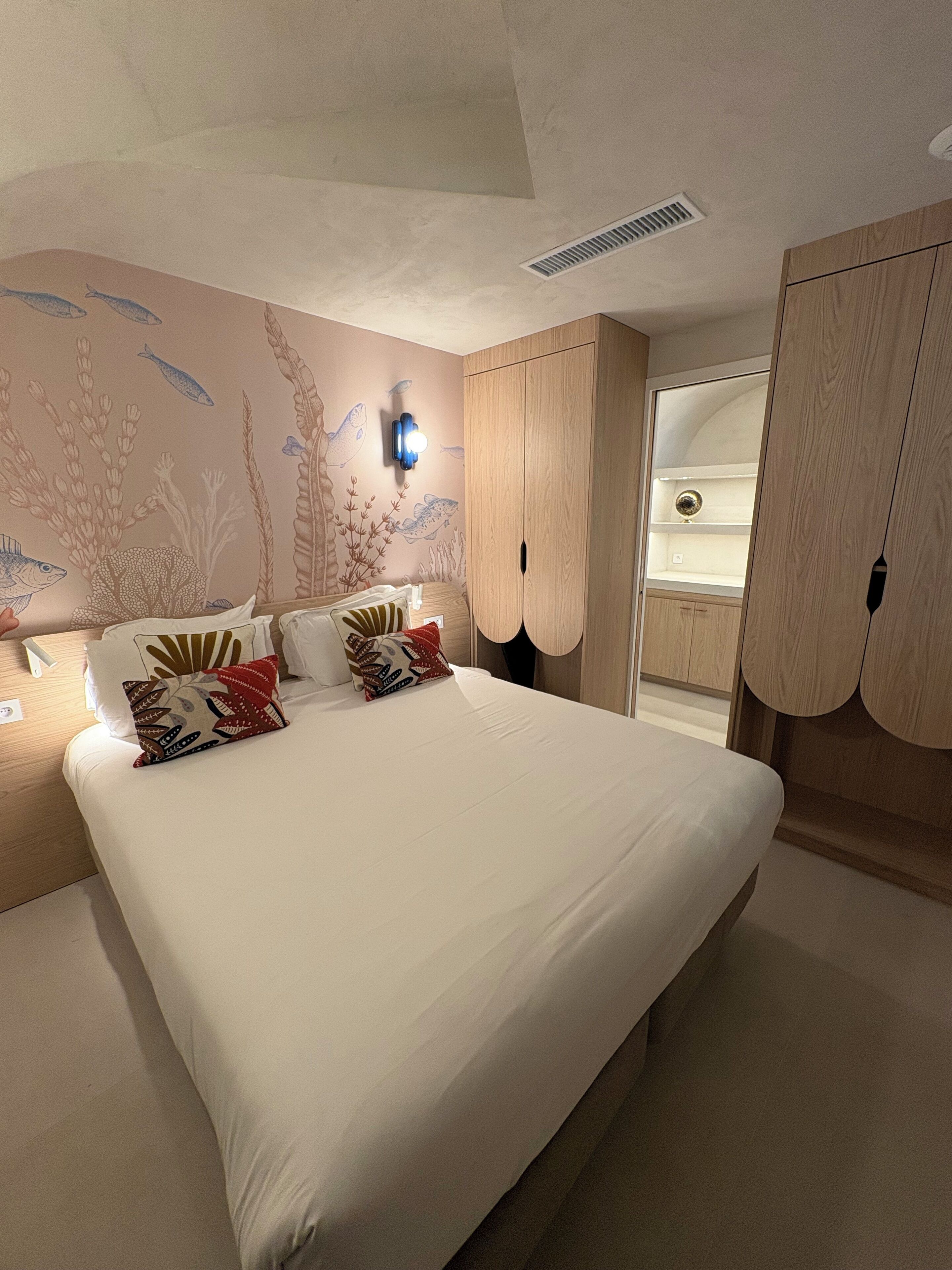 Quarto com cama grande, mesa de cabeceira, painel decorativo com peixes e corais, e guarda-roupa de madeira.