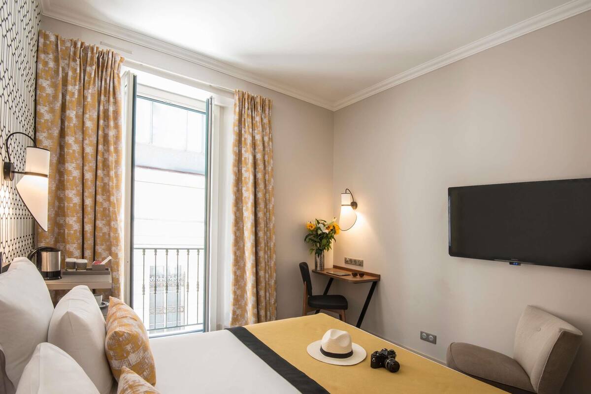 Quarto de hotel com cama, escrivaninha com luminária, cadeira, televisão e varanda com cortinas.