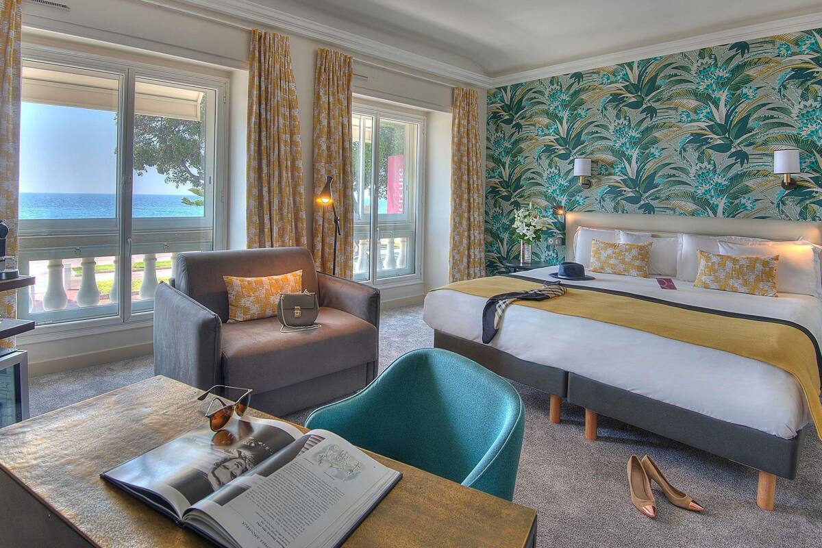 Quarto de hotel com cama, uma cadeira, uma mesa com uma revista e vista para o mar.