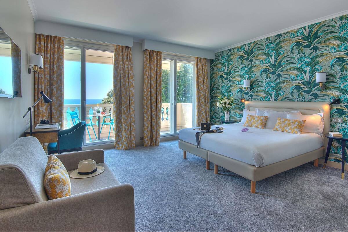 Quarto de hotel com cama, sofá, escrivaninha e vista para o exterior.