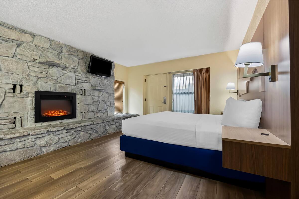 Quarto de hotel com lareira de pedra, cama com cabeceira azul, mesa de cabeceira de madeira e televisão fixada na parede.