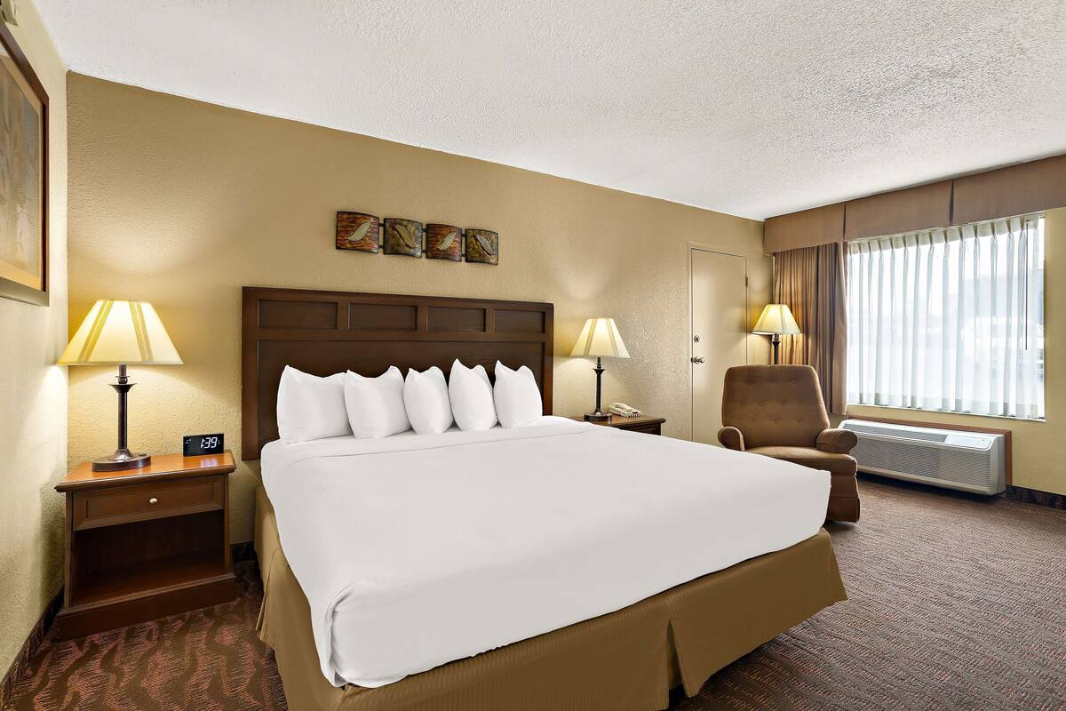 Quarto de hotel com uma cama grande, duas mesas de cabeceira com luminárias, uma cadeira e uma janela com cortinas.