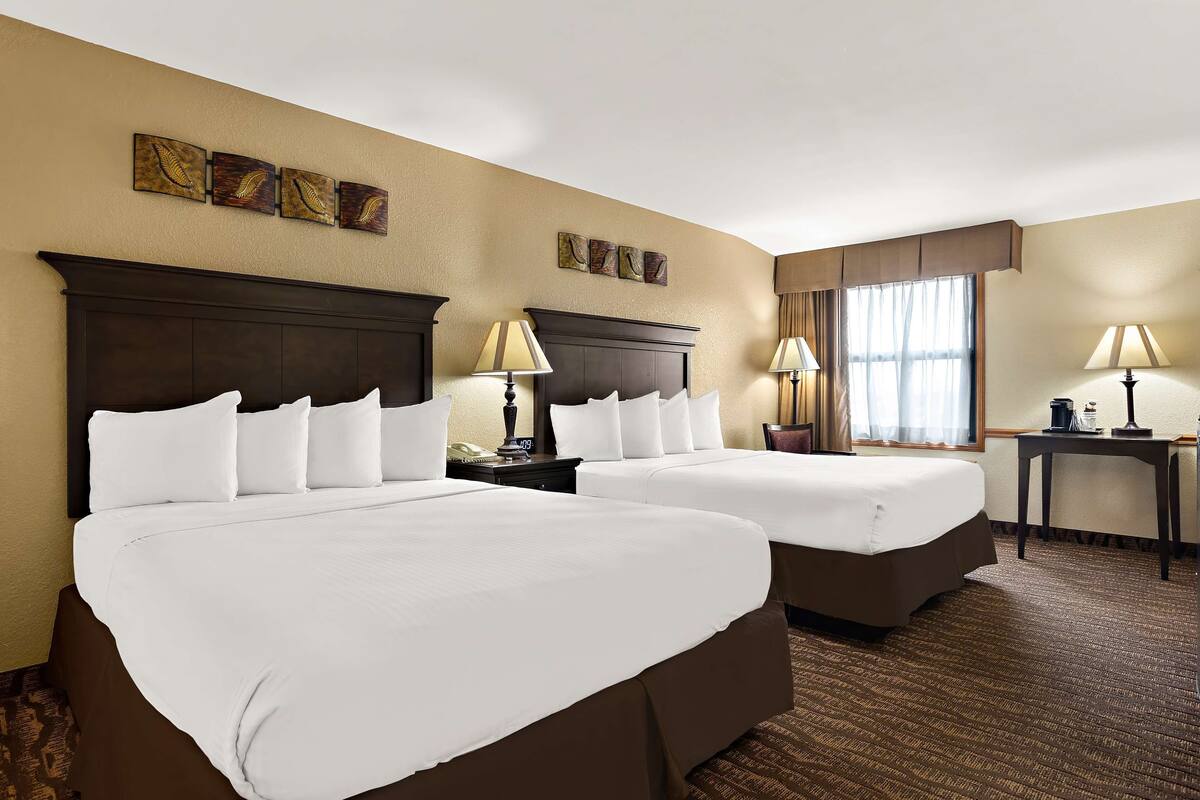 Quarto de hotel com duas camas de solteira, cabeceira de madeira, mesa de cabeceira com abajur, janela com cortinas e uma obra de arte na parede.