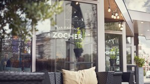 Uma área de estar externa com móveis de vime e um almofada, em frente a um edifício com a sinalização 'ZOCHER BAR & KITCHEN'.
