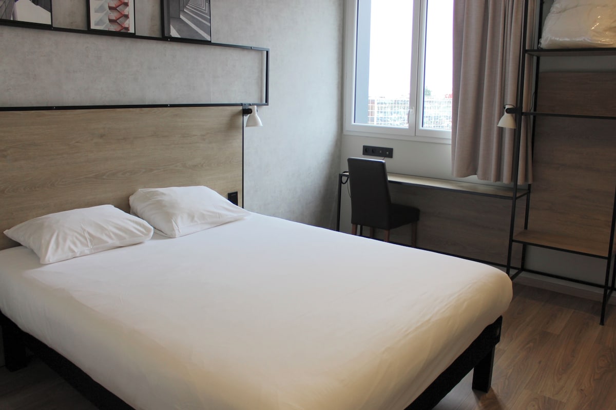 Uma cama bem arrumada com lençóis brancos em quarto moderno, com cabeceira de madeira, escrivaninha e janela com cortinas.
