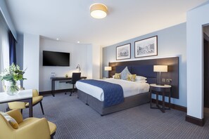 Quarto de hotel moderno com uma cama grande, uma mesa, uma cadeira, uma televisão e quadros nas paredes.