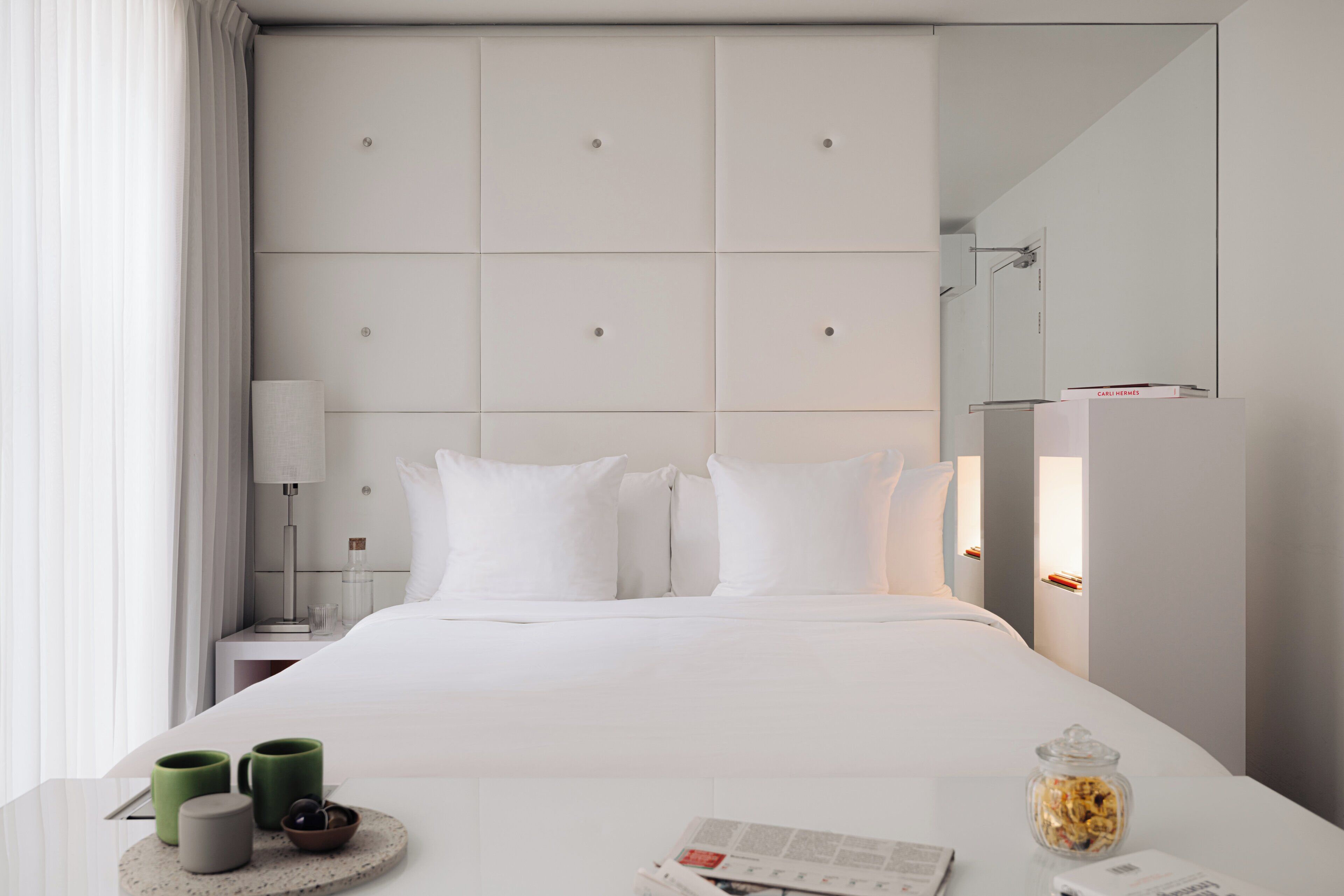 Quarto de hotel moderno com uma cama bem arrumada, uma mesa de cabeceira com abajur e uma mesinha com tigela de cereal e jornal.