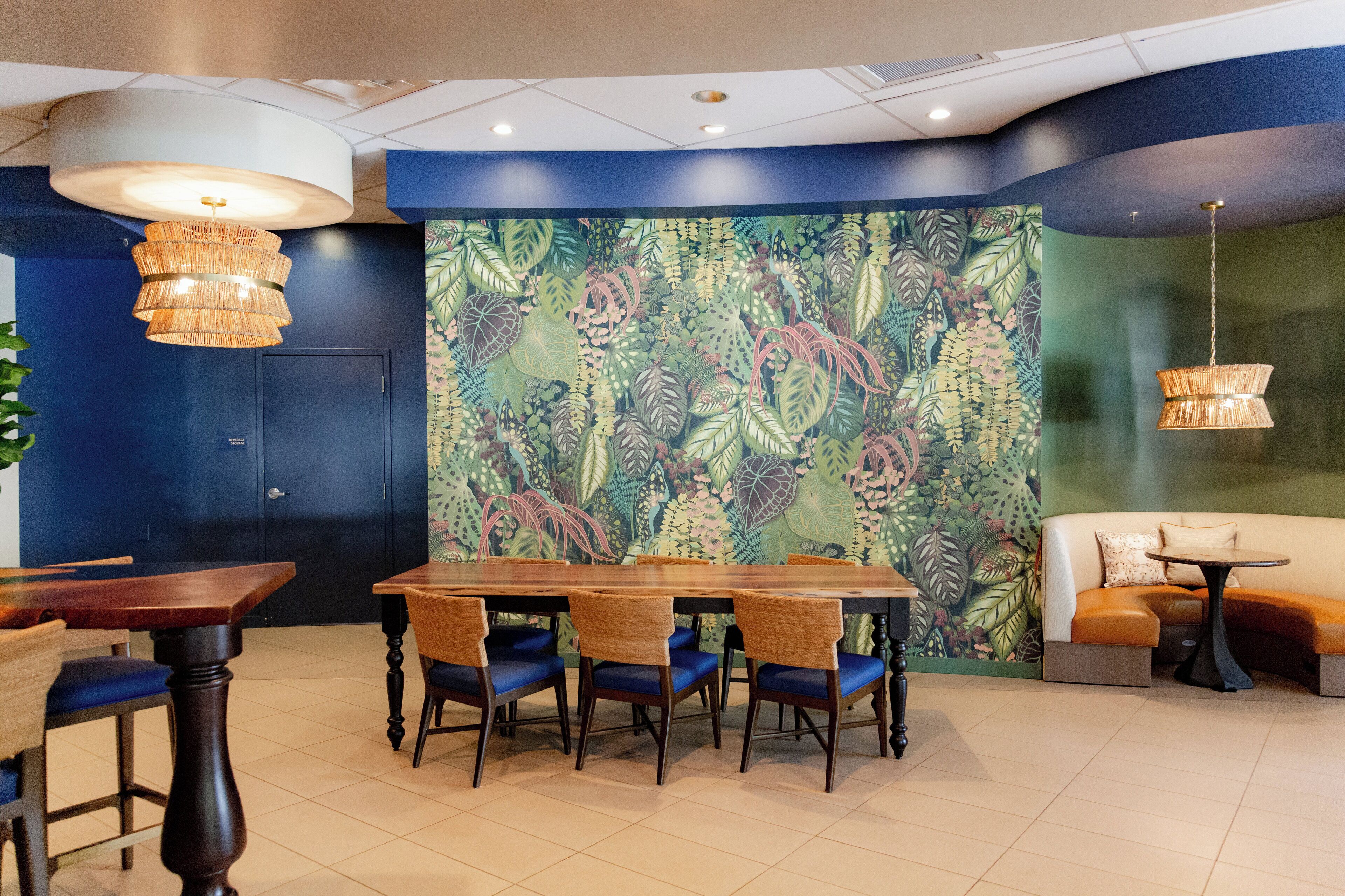 Uma sala de jantar com uma longa mesa de madeira, cadeiras estofadas em azul e um mural na parede com folhagem tropical.