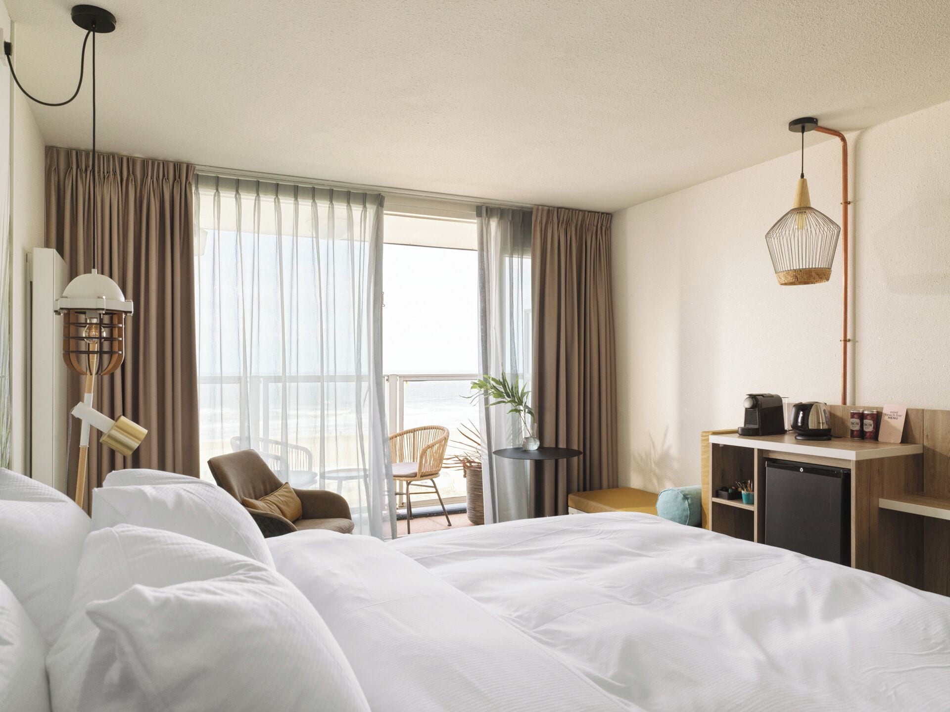 Quarto de hotel com uma cama grande, uma escrivaninha com cafeteira, uma cadeira e vista para o exterior.
