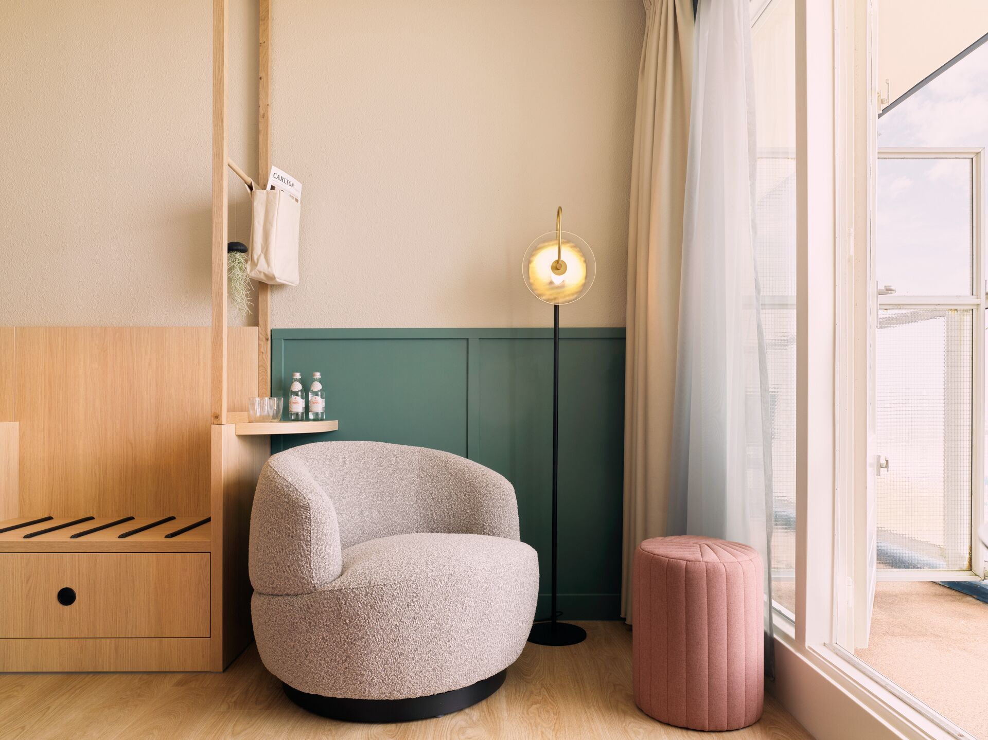 Quarto moderno com uma poltrona bege arredondada, um banquinho rosa pequeno, um abajur, uma estante com garrafas e vista pela janela.