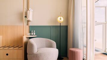 Quarto moderno com uma poltrona bege arredondada, um banquinho rosa pequeno, um abajur, uma estante com garrafas e vista pela janela.