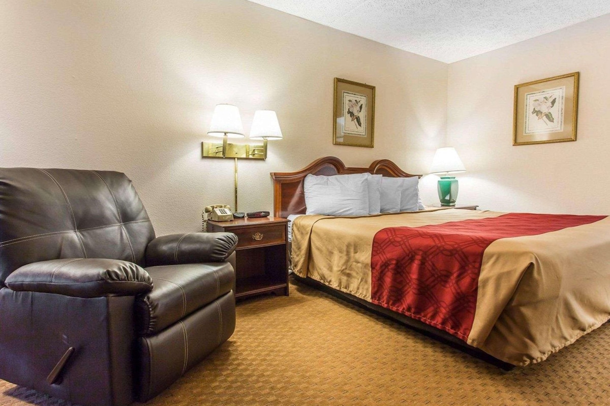 Quarto de hotel com uma poltrona de couro, cama com colcha vermelha e bege, duas luminárias de parede, mesa de cabeceira com telefone e duas fotografias emolduradas na parede.